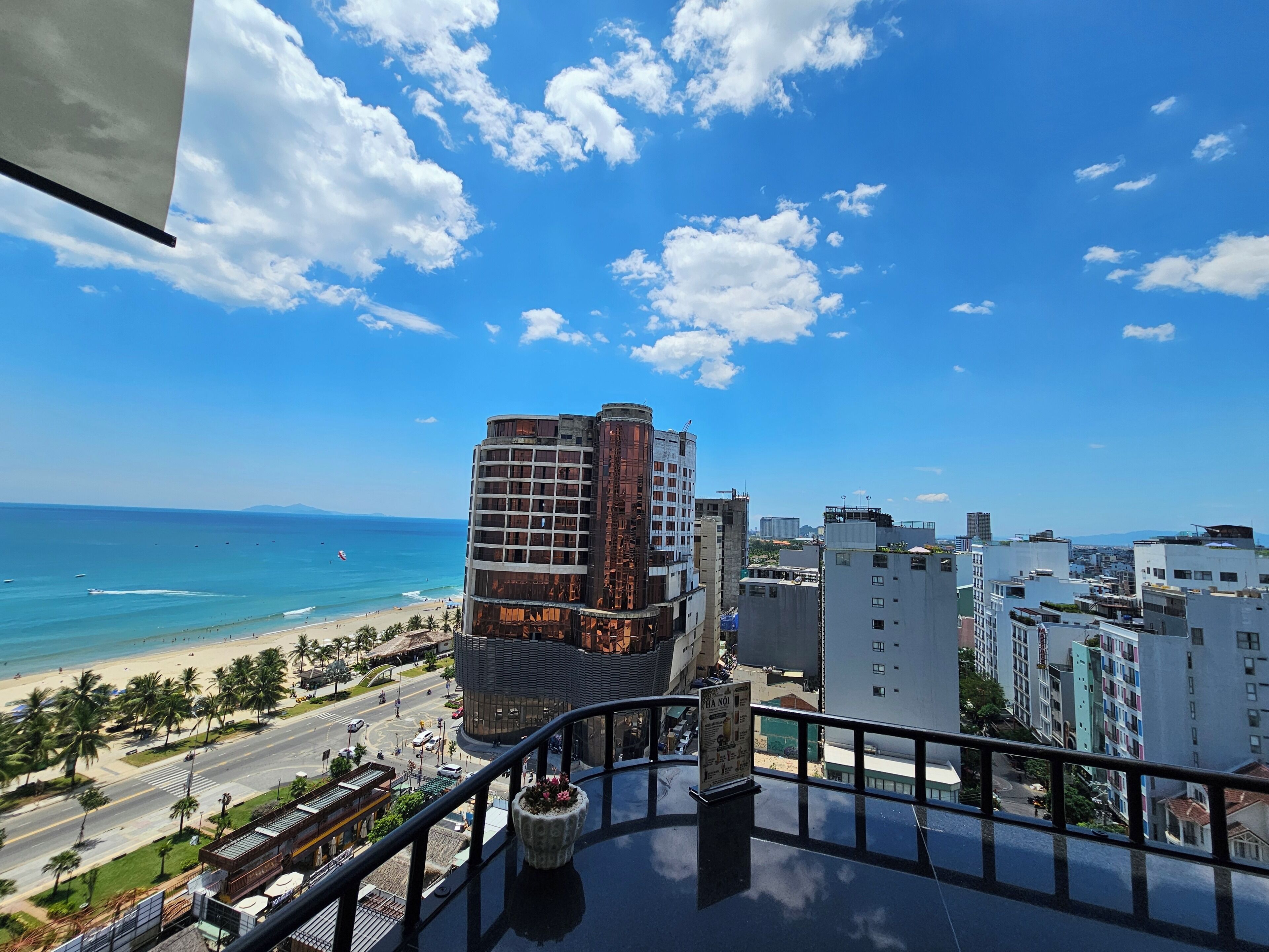 Varanda com vista para a praia e paisagem urbana, incluindo um edifício alto e uma estrada com palmeiras.