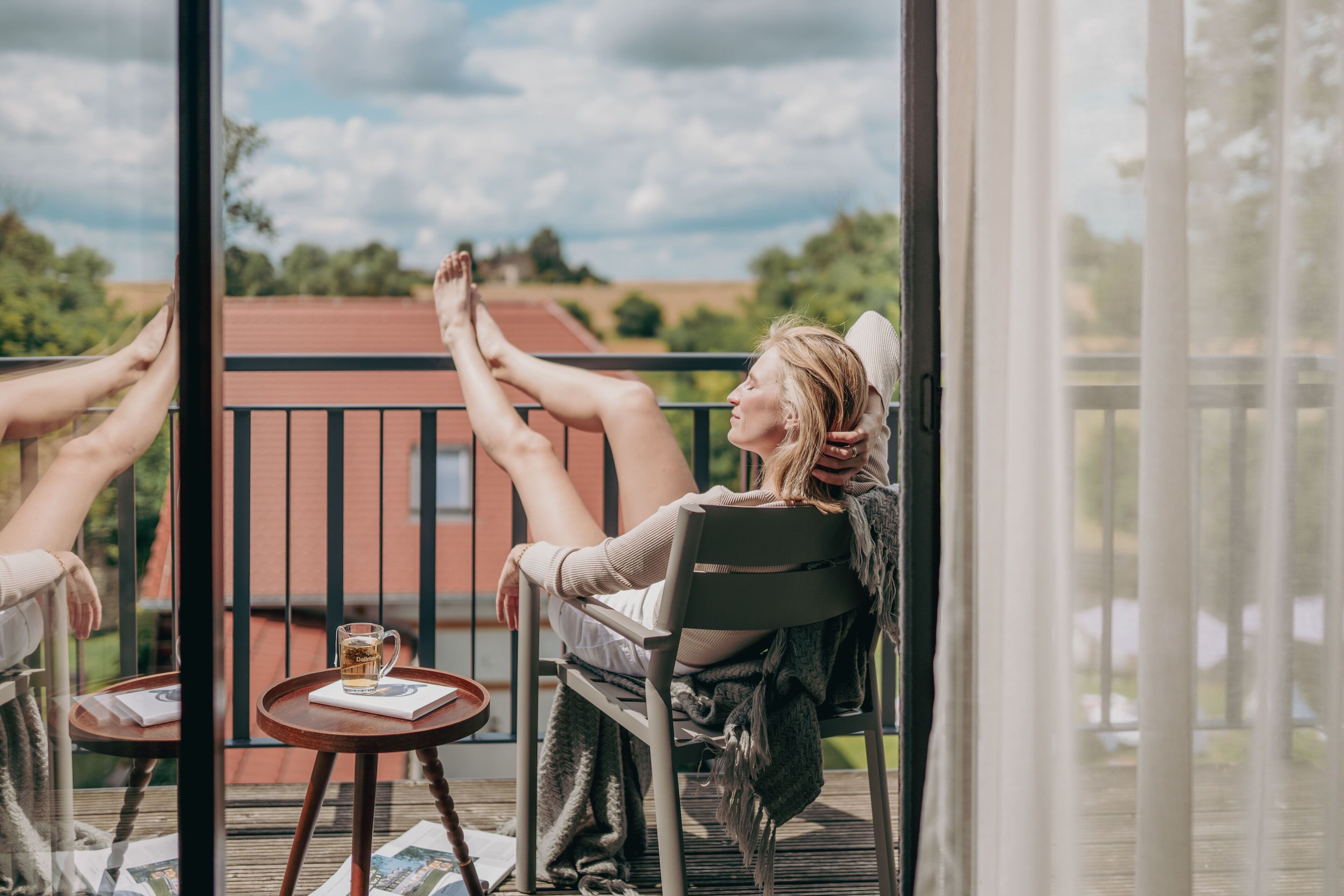 Uma pessoa sentada em uma cadeira de varanda, com as pernas estendidas, uma cerveja em um apoio e um livro sobre uma mesa pequena.
