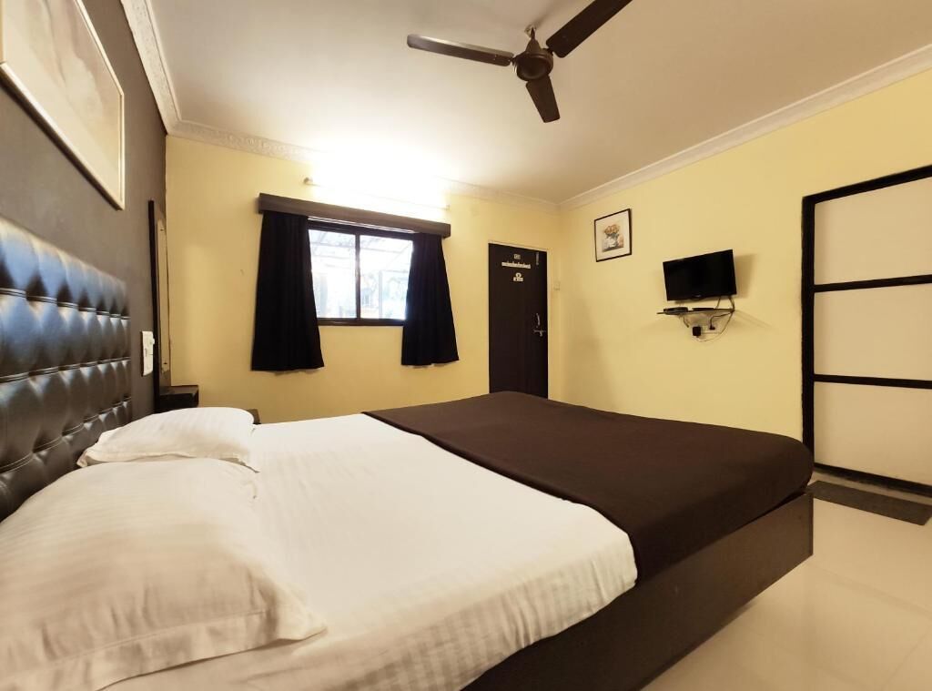 Quarto de hotel com cama, ventilador de teto, janela com cortinas, televisão e porta.