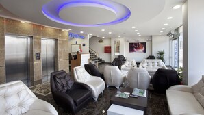 Uma área de recepção de hotel com design moderno, apresentando um balcão de atendimento, área de estar com poltronas de couro, uma mesa de centro e uma escadaria que leva aos andares superiores.