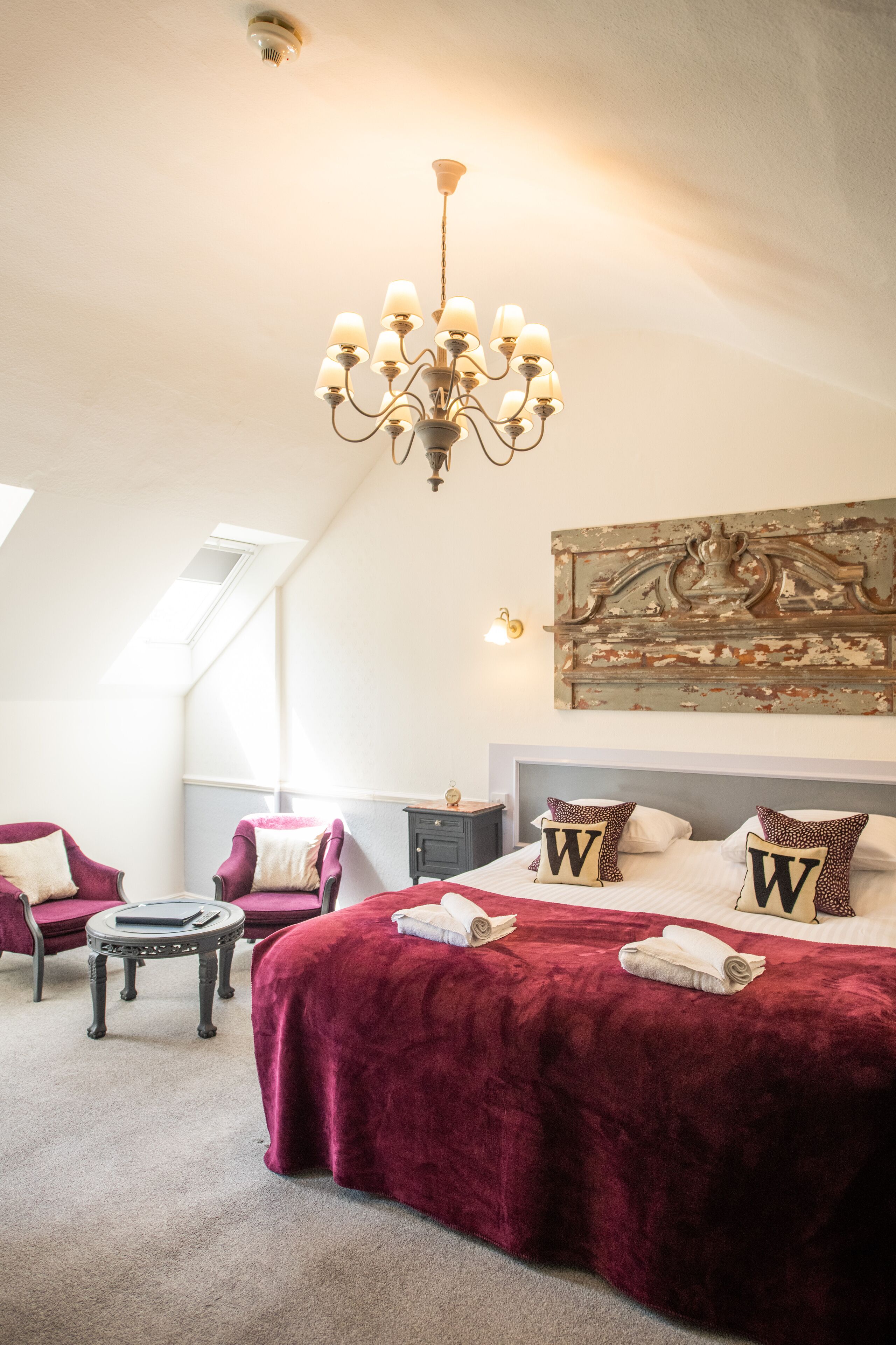 Quarto com cama grande, duas poltronas, um lustre, claraboia e um objeto decorativo na parede.