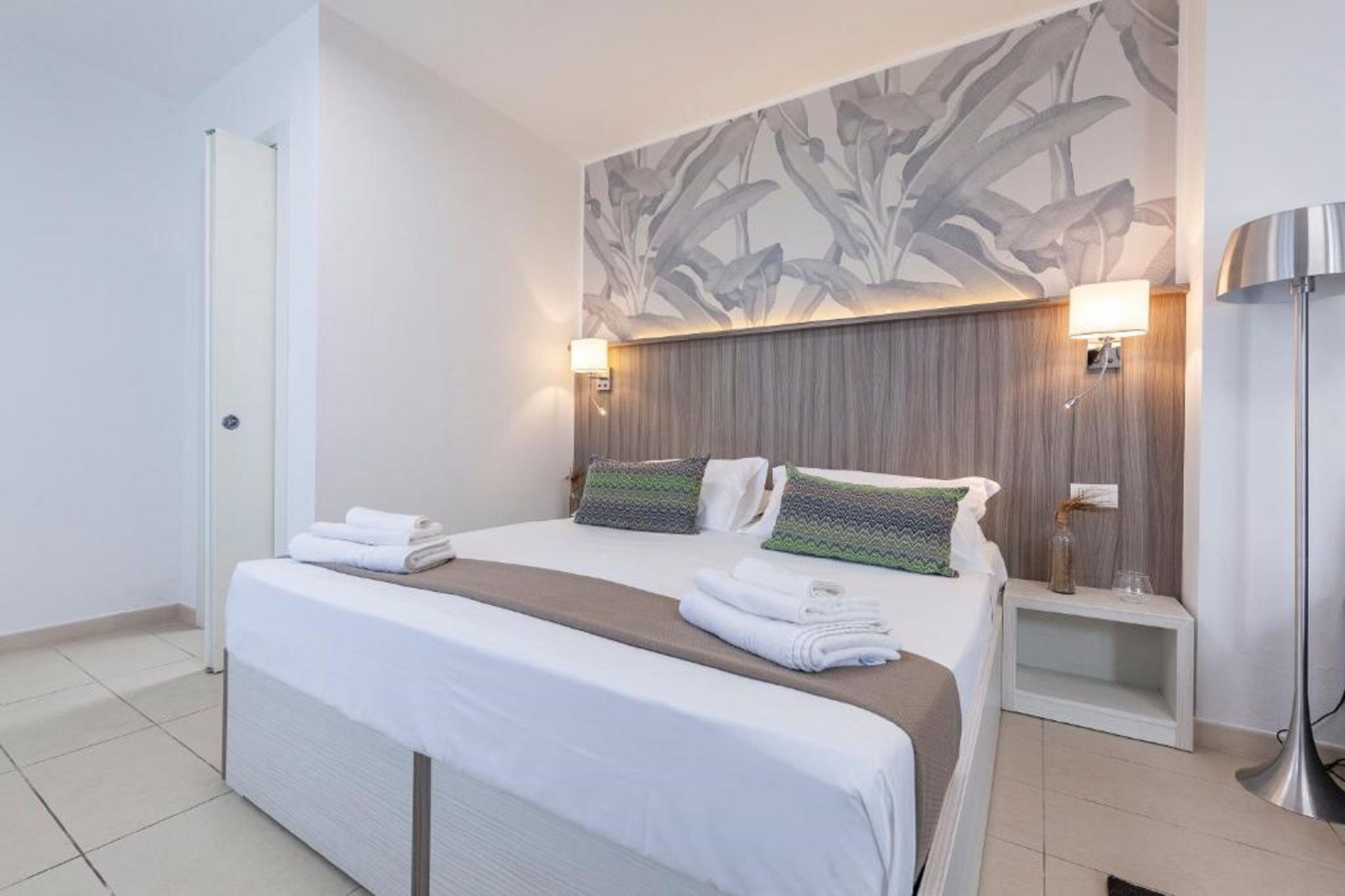 Quarto de hotel com uma cama, duas luminárias fixadas na parede, um mural decorativo e uma mesa de cabeceira com abajur.