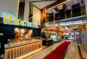 Interior moderno de hotel com um destaque para a placa 'UESPIA HOTEL', um balcão de exposição com sobremesas e uma exposição de carros antigos.