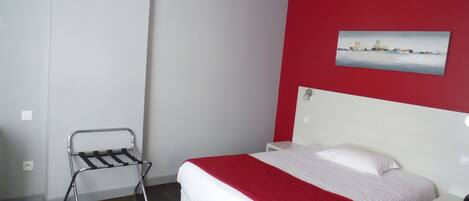 Quarto de hotel com cama, mesa de cabeceira, parede vermelha e uma pintura de paisagem urbana.