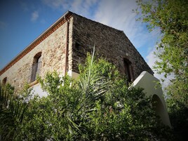 Um edifício de pedra com janelas em arco e varanda, rodeado por vegetação exuberante.