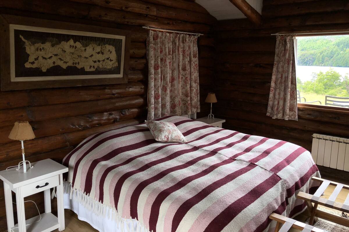 Quarto de cabana de madeira com cama, mesa de cabeceira, luminária e janela com cortinas.