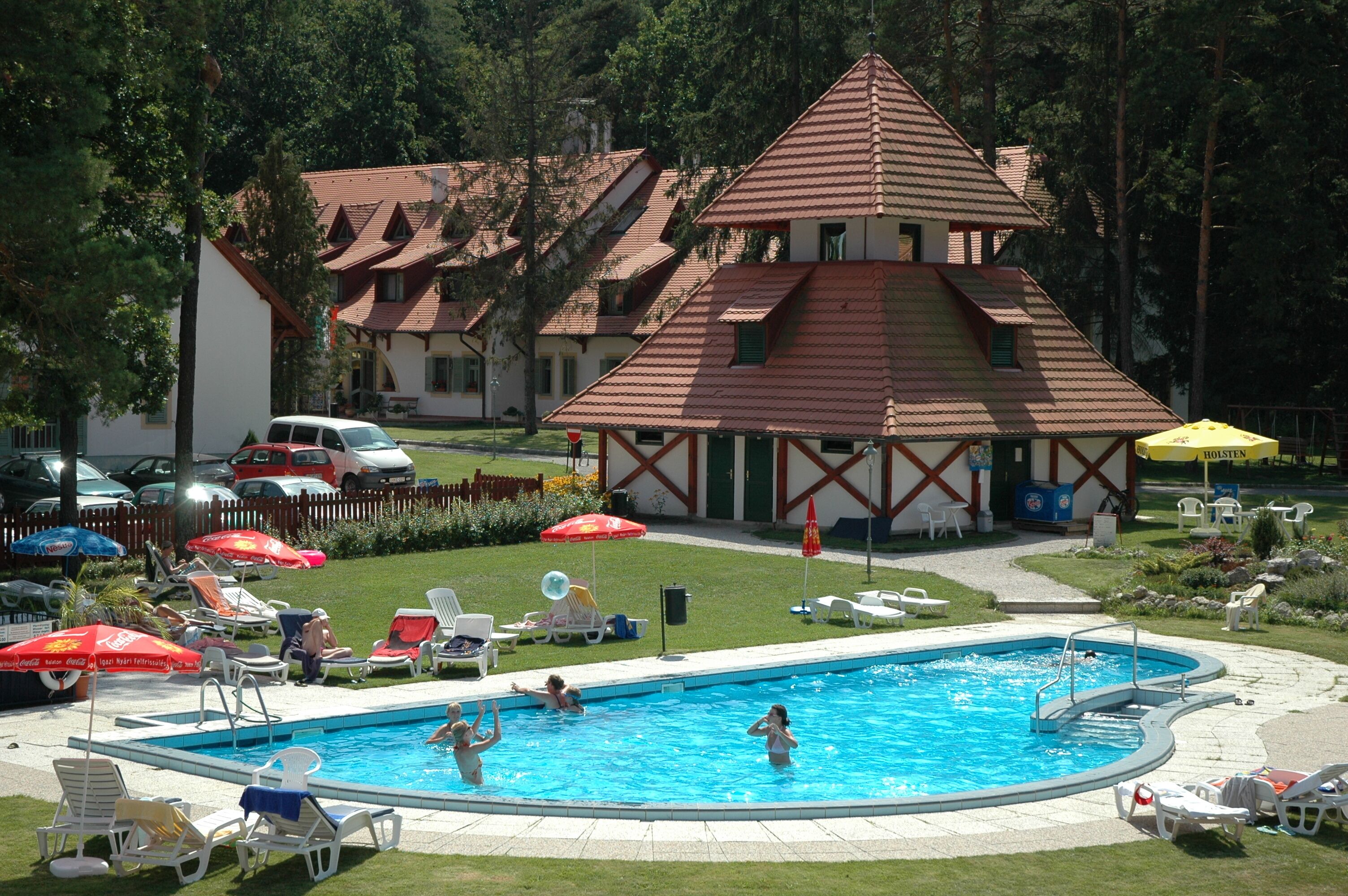 Uma área de piscina com espreguiçadeiras e guarda-sóis, rodeada por um edifício com telhado de telhas vermelhas.
