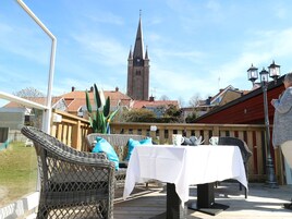 Um pátio externo com móveis de vime e uma mesa, uma torre de igreja ao fundo e um edifício com telhado vermelho.