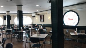 Um restaurante com várias mesas e cadeiras, uma janela grande com a placa 'Greece', e um aparelho de ar condicionado no teto.