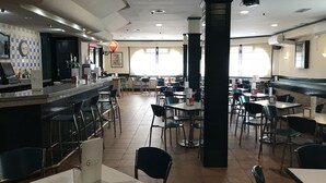 Uma cafeteria ampla com várias mesas e cadeiras, um balcão com o logotipo da Coca-Cola e uma grande janela que proporciona iluminação natural.