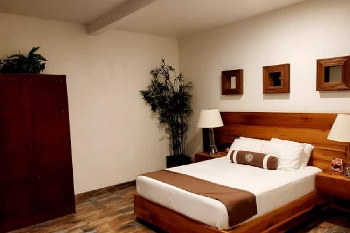 Quarto com piso de madeira, cama com roupa de cama branca, mesa de cabeceira, luminária e espelho na parede.