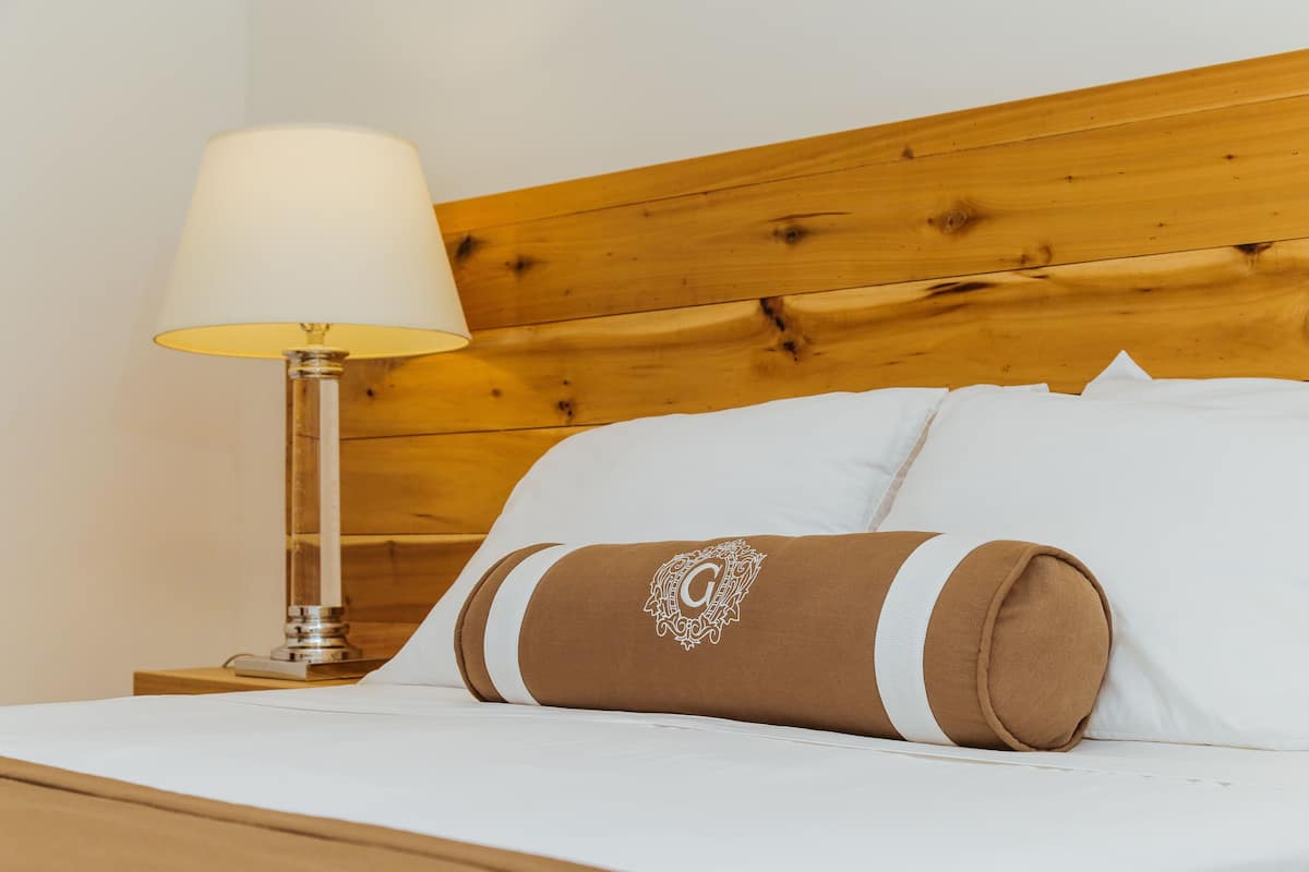 Cama bem arrumada com lençóis brancos, travesseiro marrom com monograma e abajur sobre mesa de cabeceira de madeira.