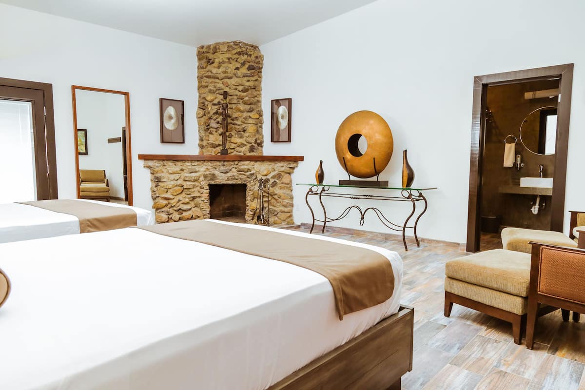 Quarto de hotel com duas camas, uma lareira de pedra, um espelho redondo e uma porta de madeira.