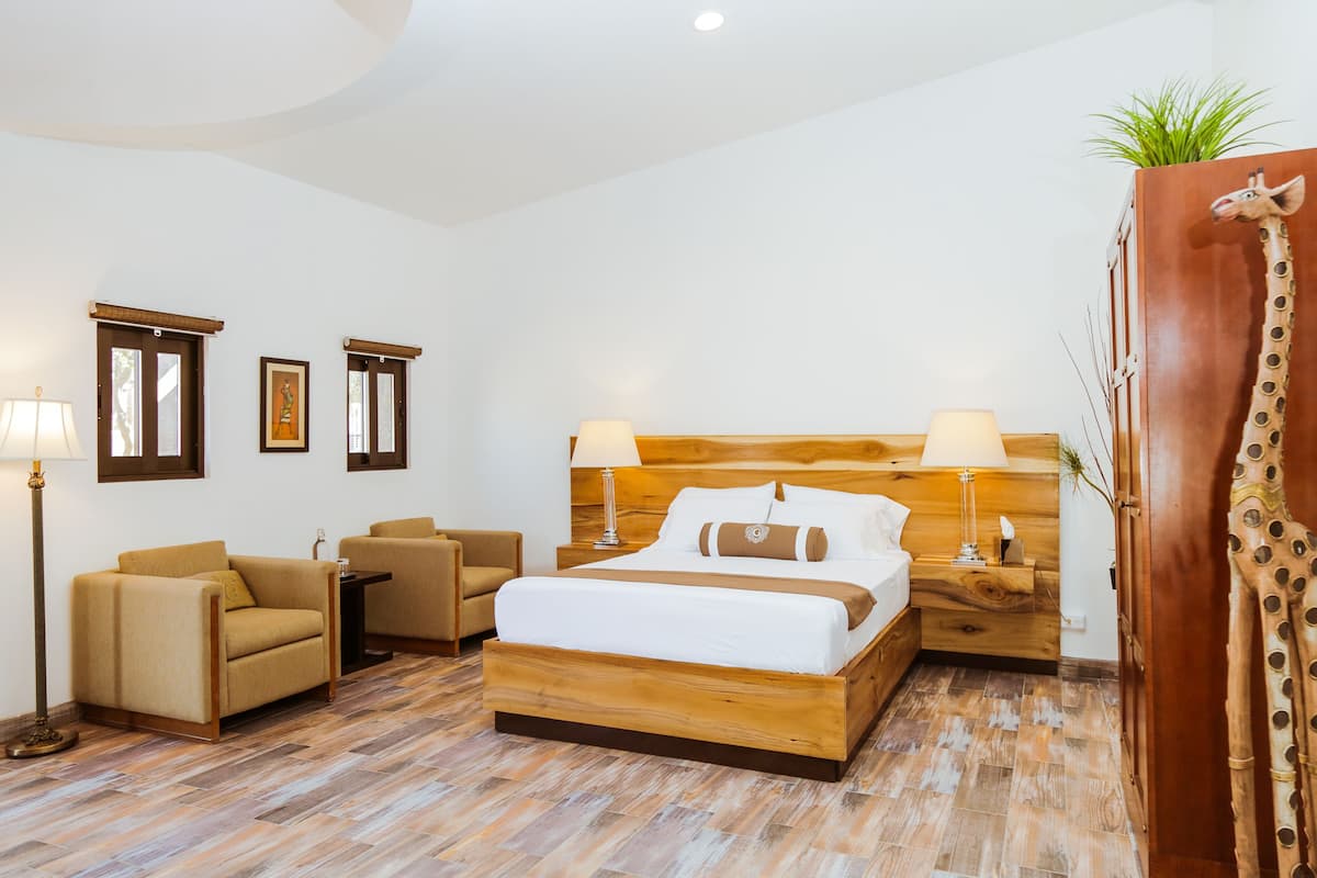 Quarto de hotel com cama de madeira, duas poltronas, mesa de cabeceira e uma luminária.