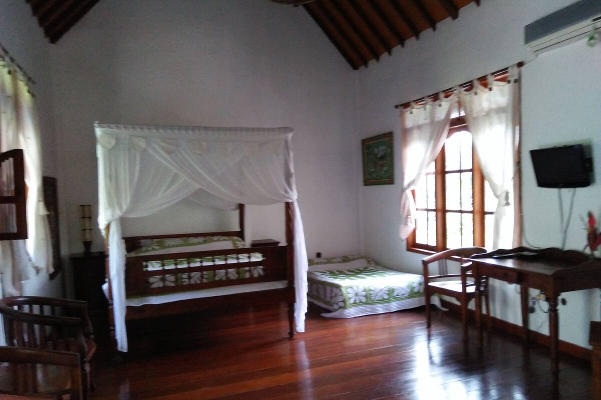 Um quarto com piso de madeira, cama com dossel, escrivaninha, cadeiras e televisão na parede.