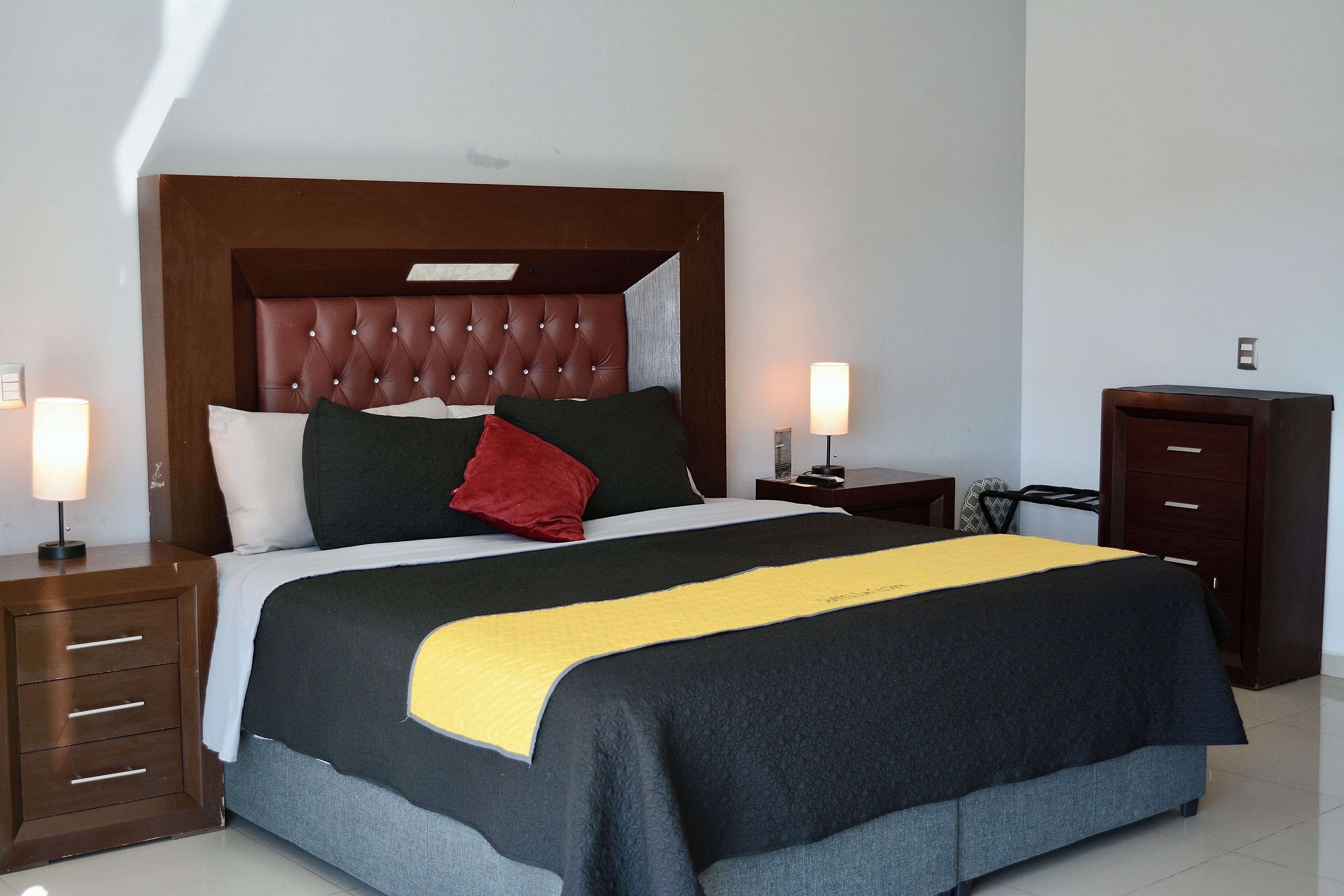 Cama bem arrumada com cabeceira estofada, duas mesas de cabeceira com abajures e uma cômoda de madeira.