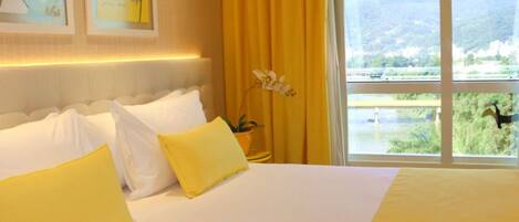 Quarto de hotel com uma cama grande, almofadas amarelas, um vaso de planta e vista para as montanhas pela janela.