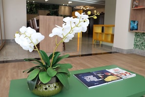 Uma mesa de recepção com superfície verde, um vaso com orquídeas brancas e uma revista.