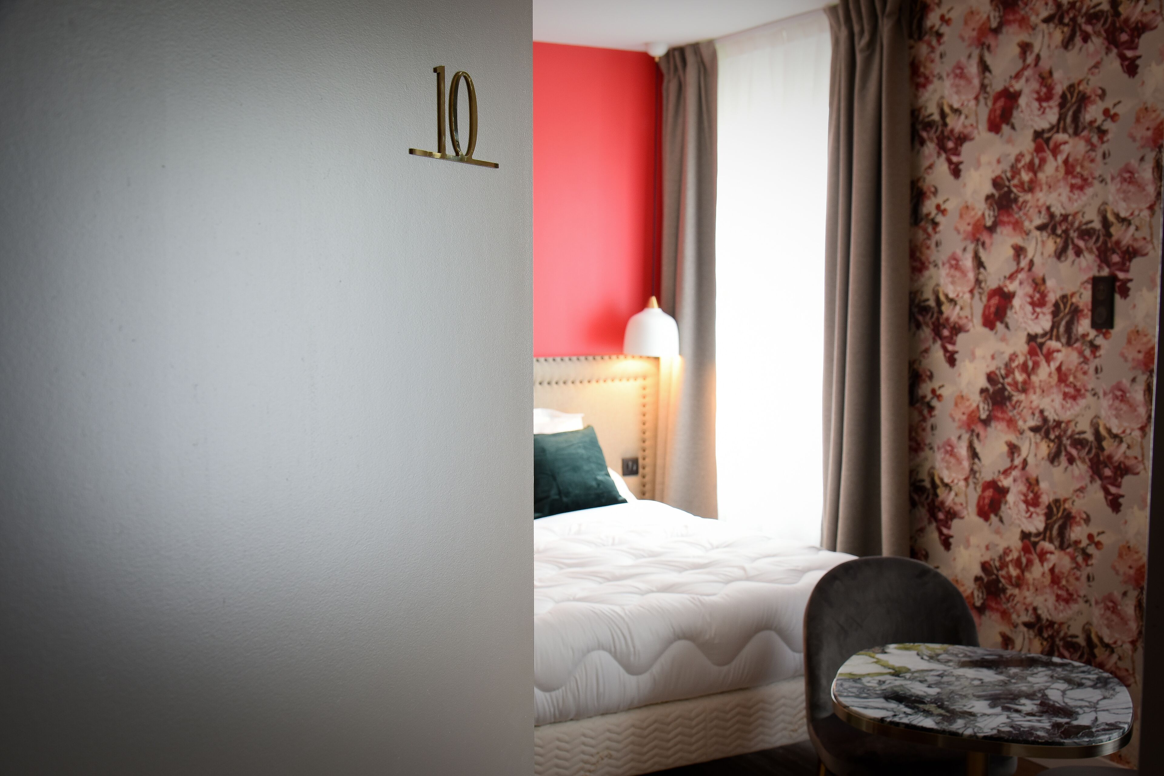 Quarto de hotel com cama, cadeira, mesinha, abajur e papel de parede floral.