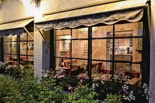 Um restaurante com mesas ao ar livre, grandes janelas e interior bem iluminado.