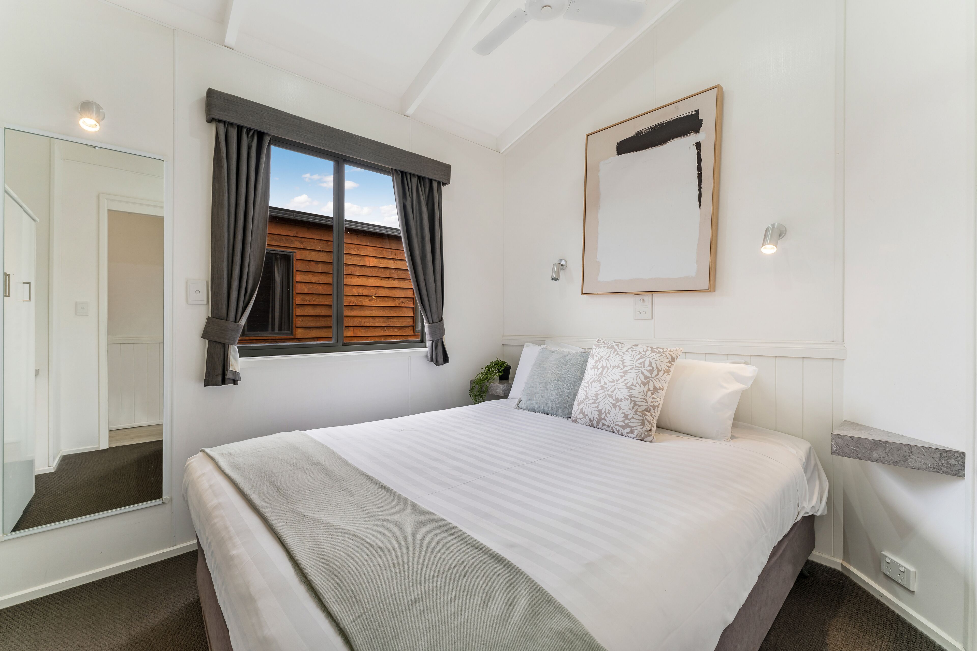 Quarto com uma cama grande, janela com cortinas, espelho na parede e uma pequena planta no criado-mudo.