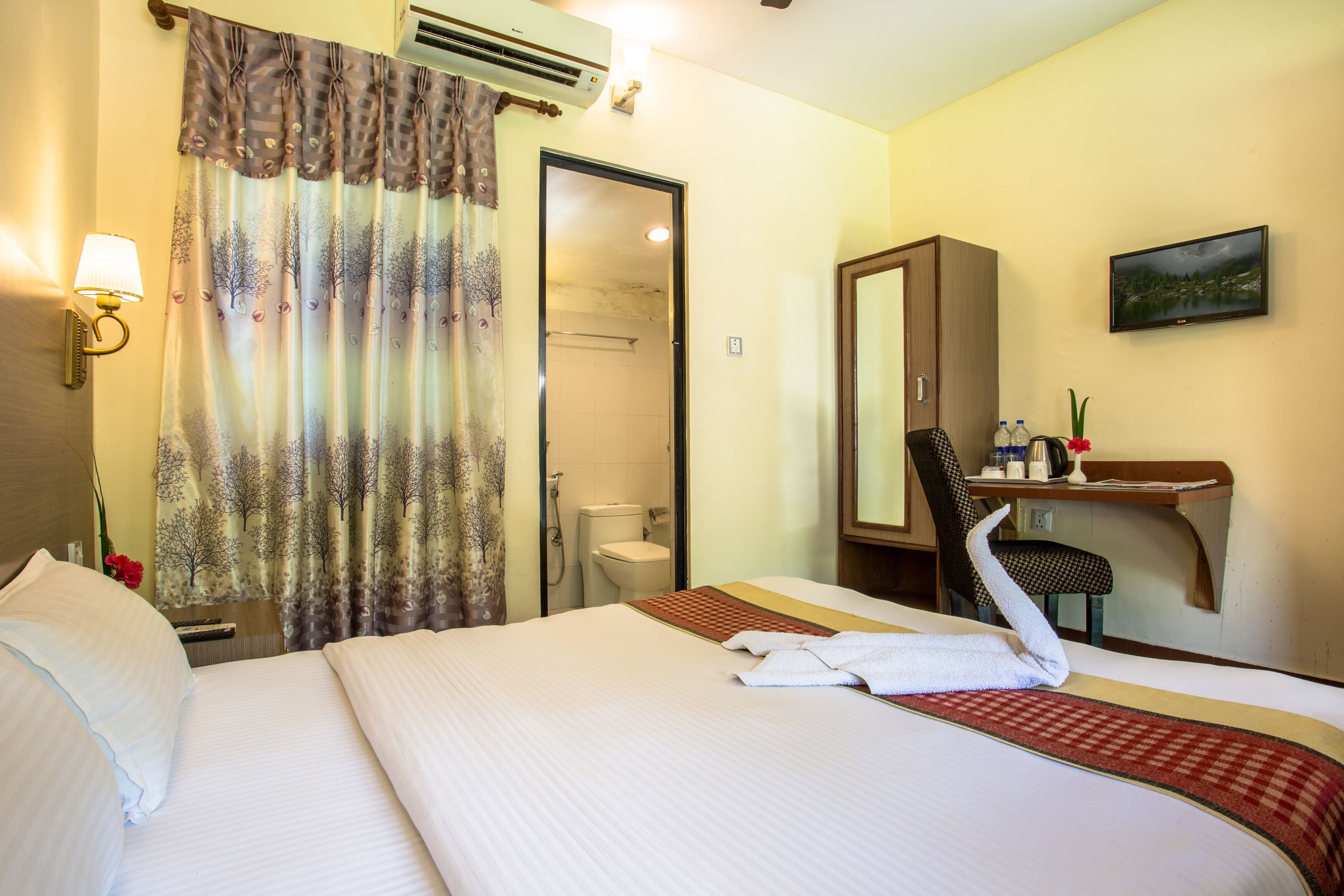 Quarto de hotel com cama, escrivaninha, cadeira, espelho e área de chuveiro.
