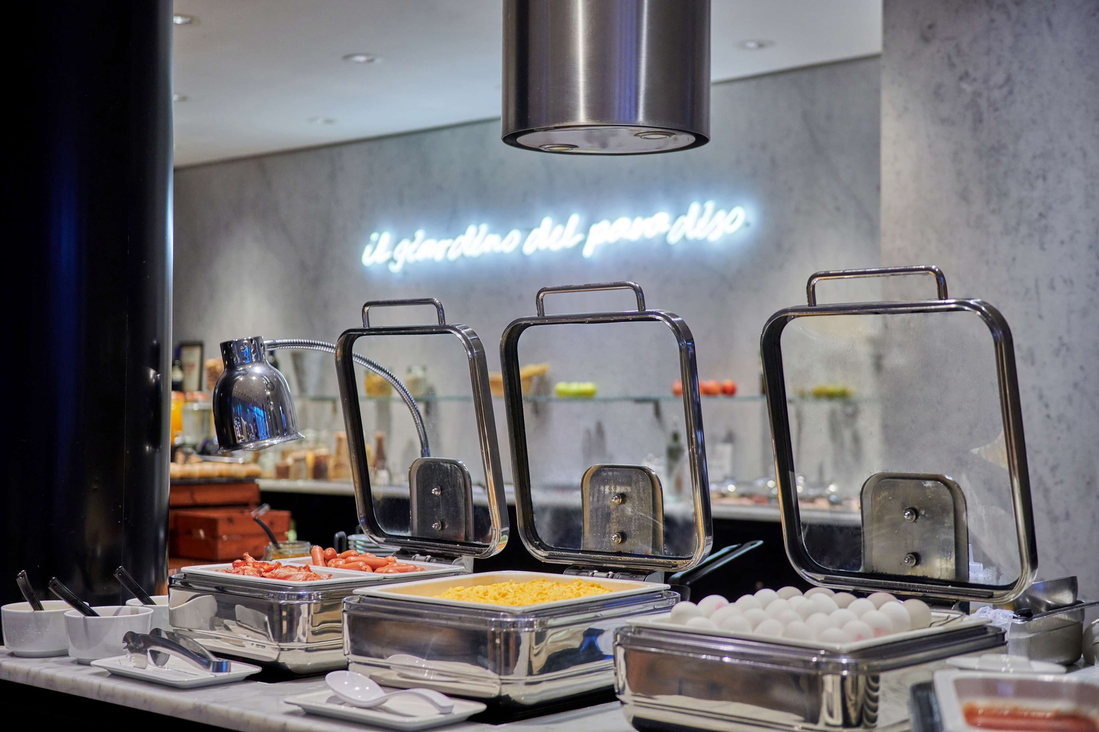 Buffet montado com diversos alimentos em recipientes transparentes, um letreiro neon ao fundo e uma coifa moderna acima.