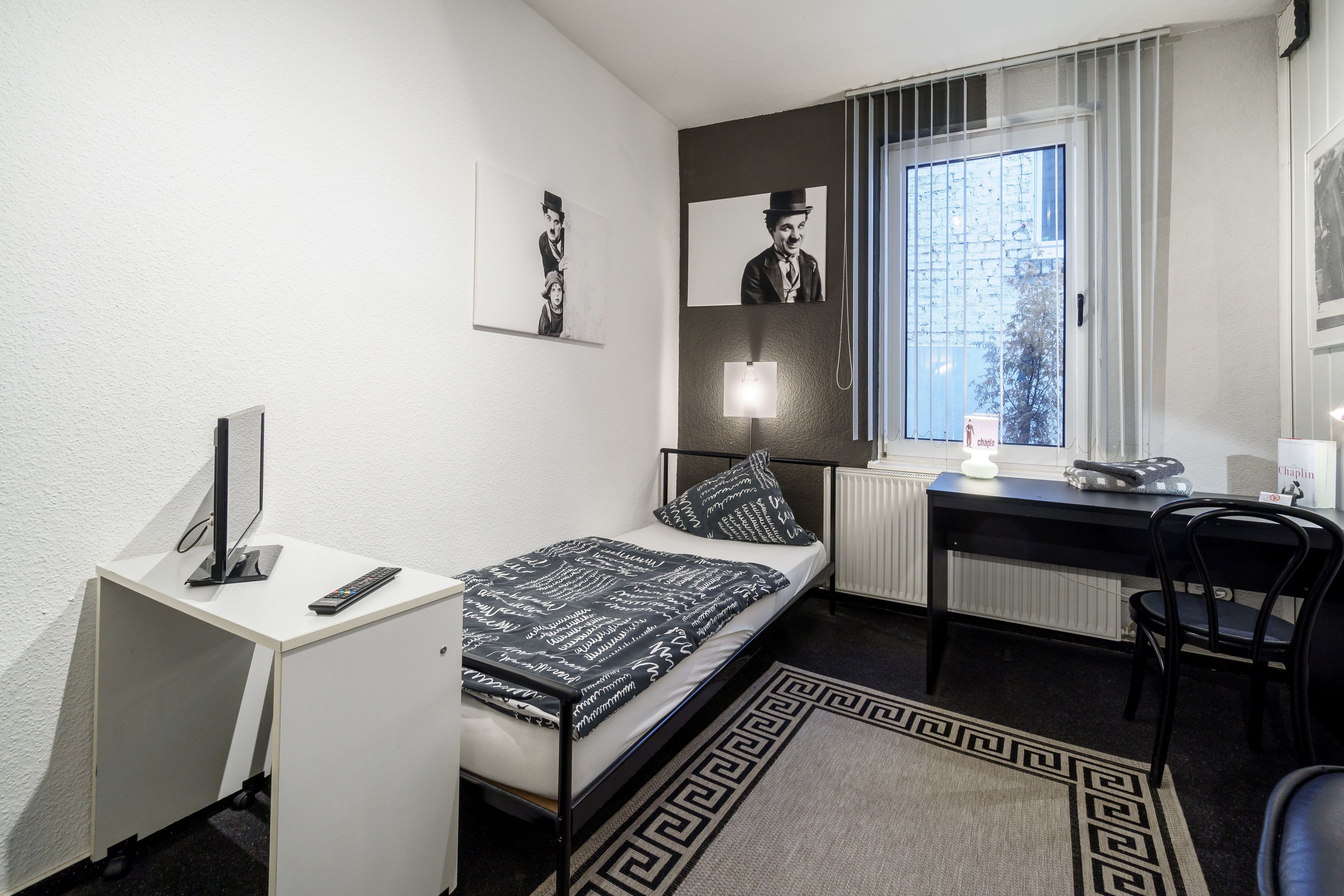 Um quarto com cama, escrivaninha, cadeira, radiador e janela com cortinas.