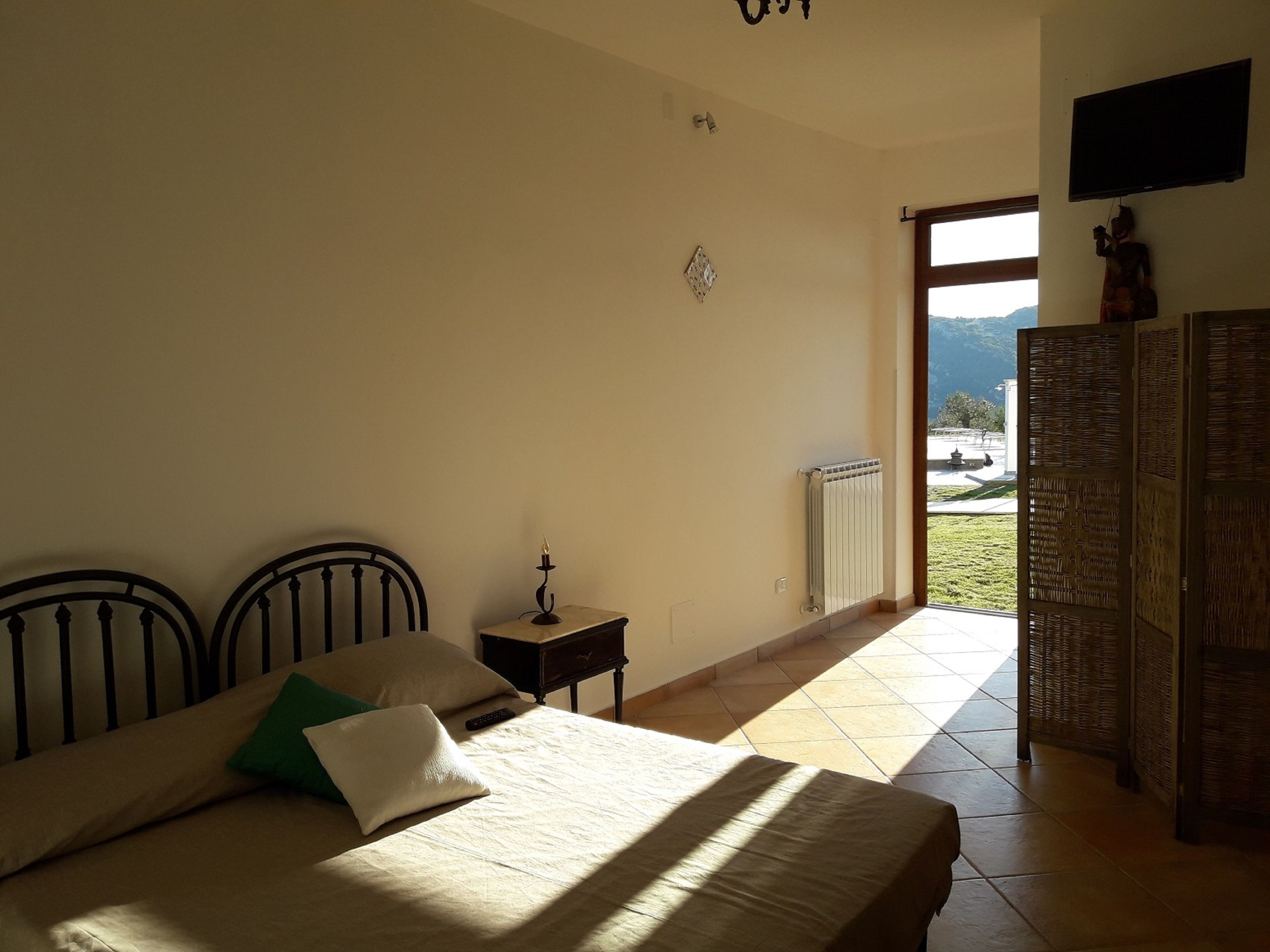 Quarto com cama, mesa de cabeceira, televisão e vista para um jardim por uma porta aberta.