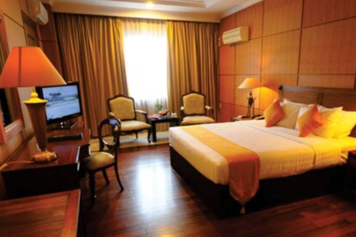 Quarto de hotel com uma cama grande, duas cadeiras, uma TV e luminárias.
