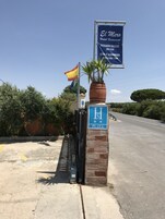 Entrada de hotel com placa de El Mero Hostal Restaurante, uma bandeira e uma planta de palmeira.