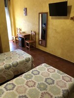 Quarto de hotel com duas camas, uma escrivaninha com cadeira, uma mesinha, televisão presa à parede e um espelho.