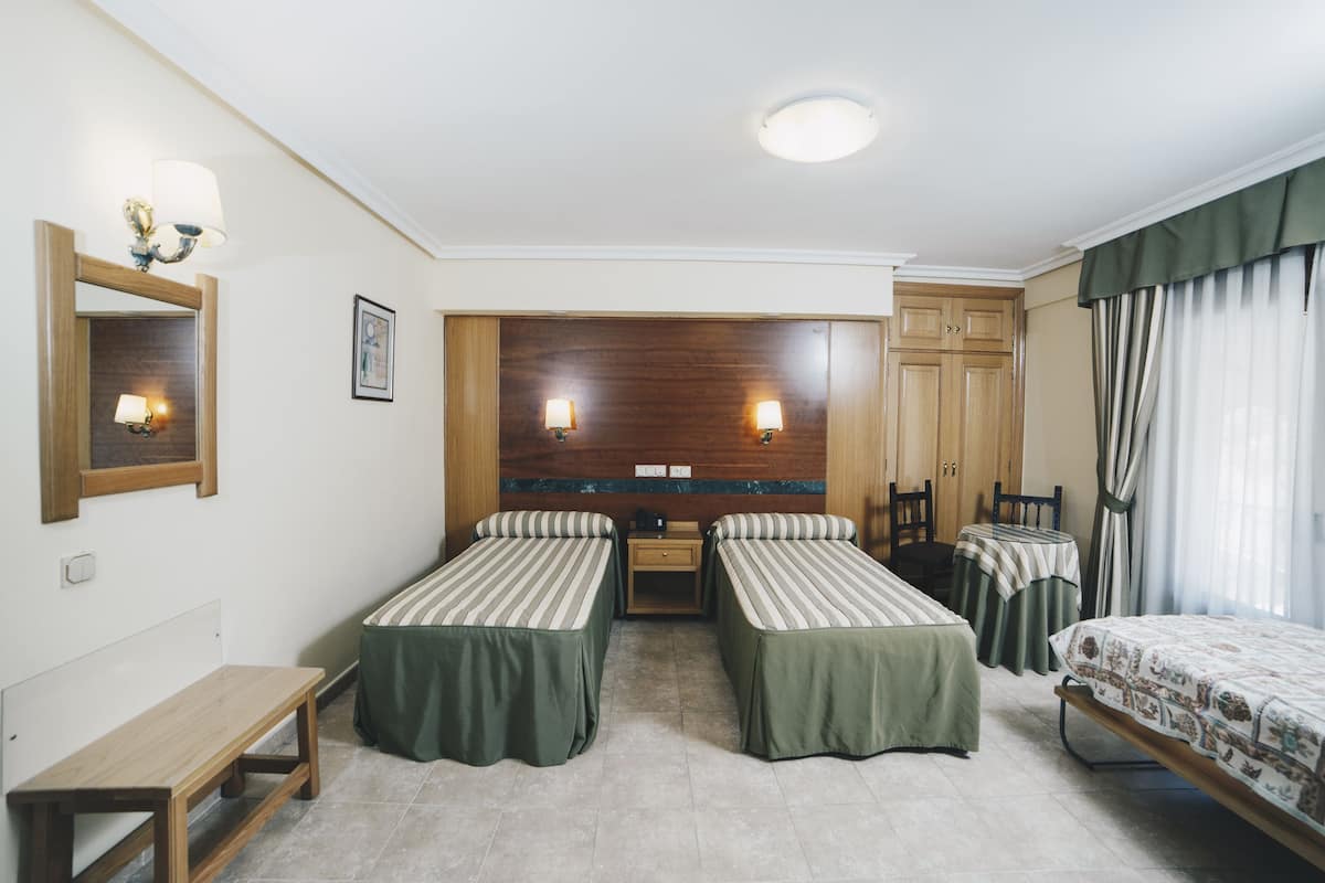 Quarto de hotel com duas camas de solteiro, cabeceira de madeira, uma mesinha, um banquinho e um espelho.