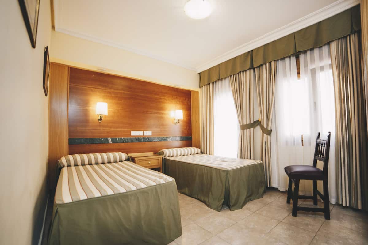 Quarto de hotel com duas camas de solteiro, cabeceira de madeira, duas luminárias de cabeceira, uma cadeira e uma janela com cortinas.