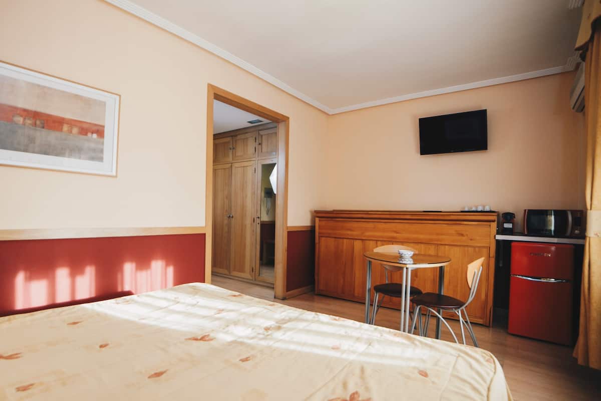 Quarto de hotel com cama, escrivaninha, cadeira, televisão e cozinha compacta.