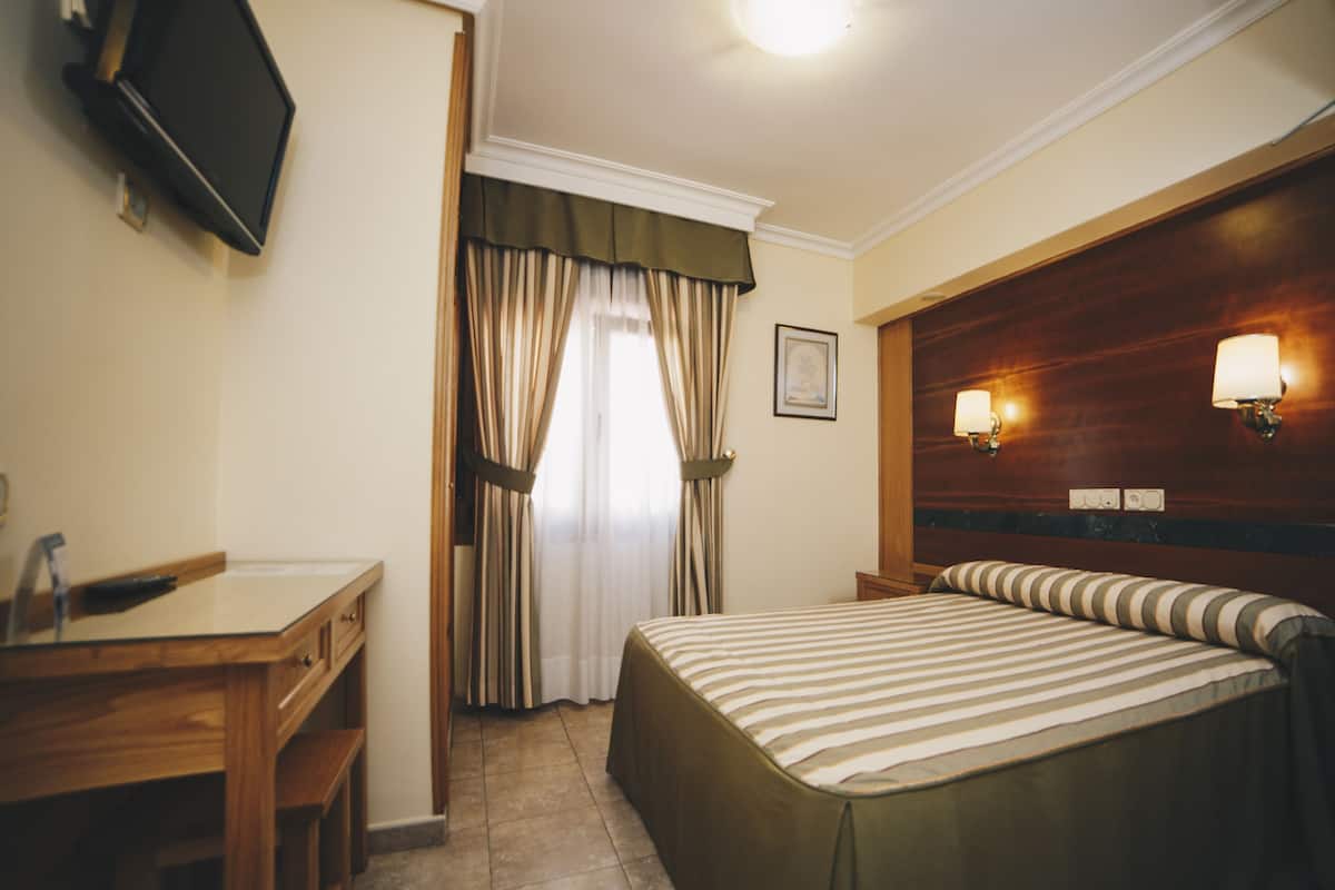 Quarto de hotel com cama, escrivaninha, televisão, janela com cortinas e luminárias fixadas na parede.
