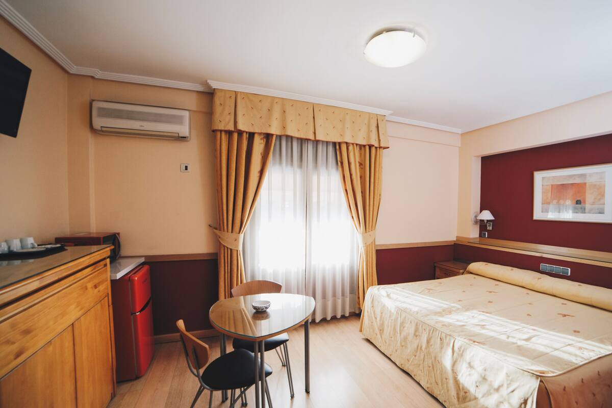 Quarto de hotel com uma cama, uma mesinha com cadeiras, uma escrivaninha com um aparelho vermelho e uma janela com cortinas.
