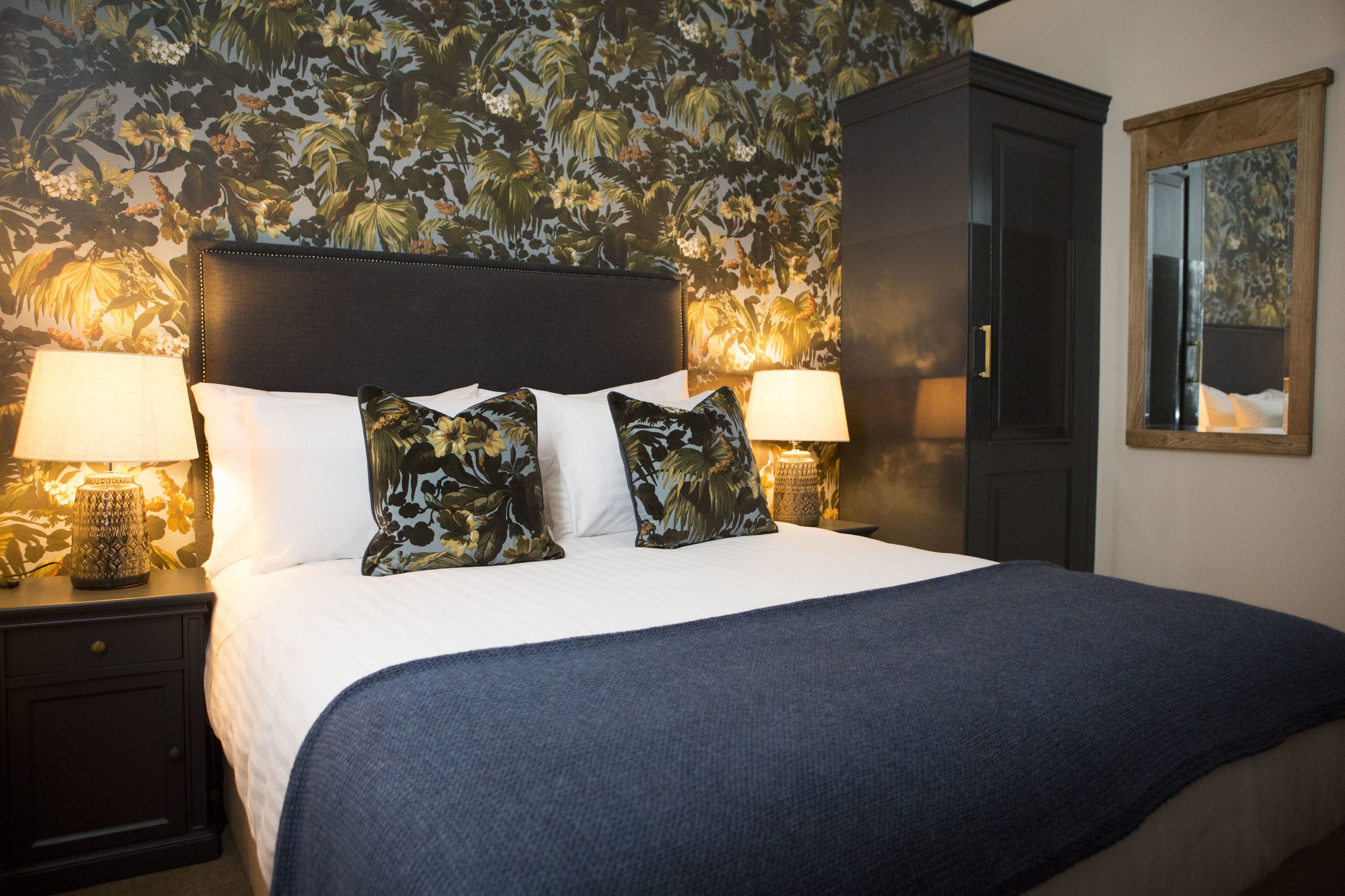 Cama bem arrumada com cabeceira escura e almofadas estampadas com flores, com duas luminárias de cabeceira em um criado-mudo.
