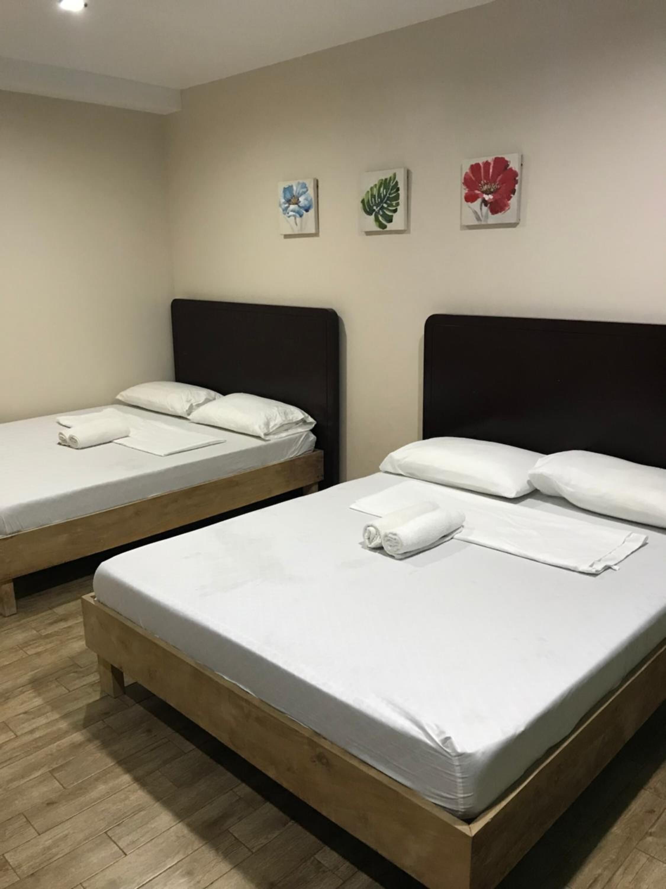 Quarto de hotel com duas camas de solteiro, cabeceiras de madeira, roupa de cama branca e três peças de arte floral na parede.
