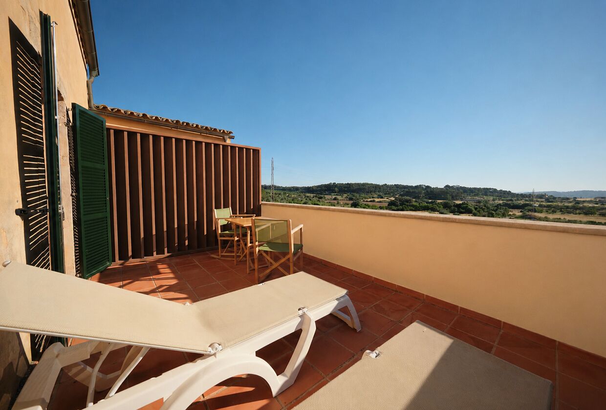 Uma varanda com mesa e cadeiras, vista para o campo e um edifício com telhado de telhas.