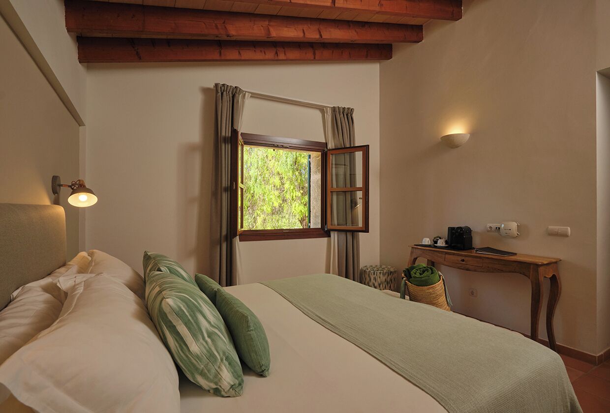 Quarto com cama, janela com cortinas, abajur na mesa de cabeceira, pequena mesa com luminária e iluminação embutida na parede.