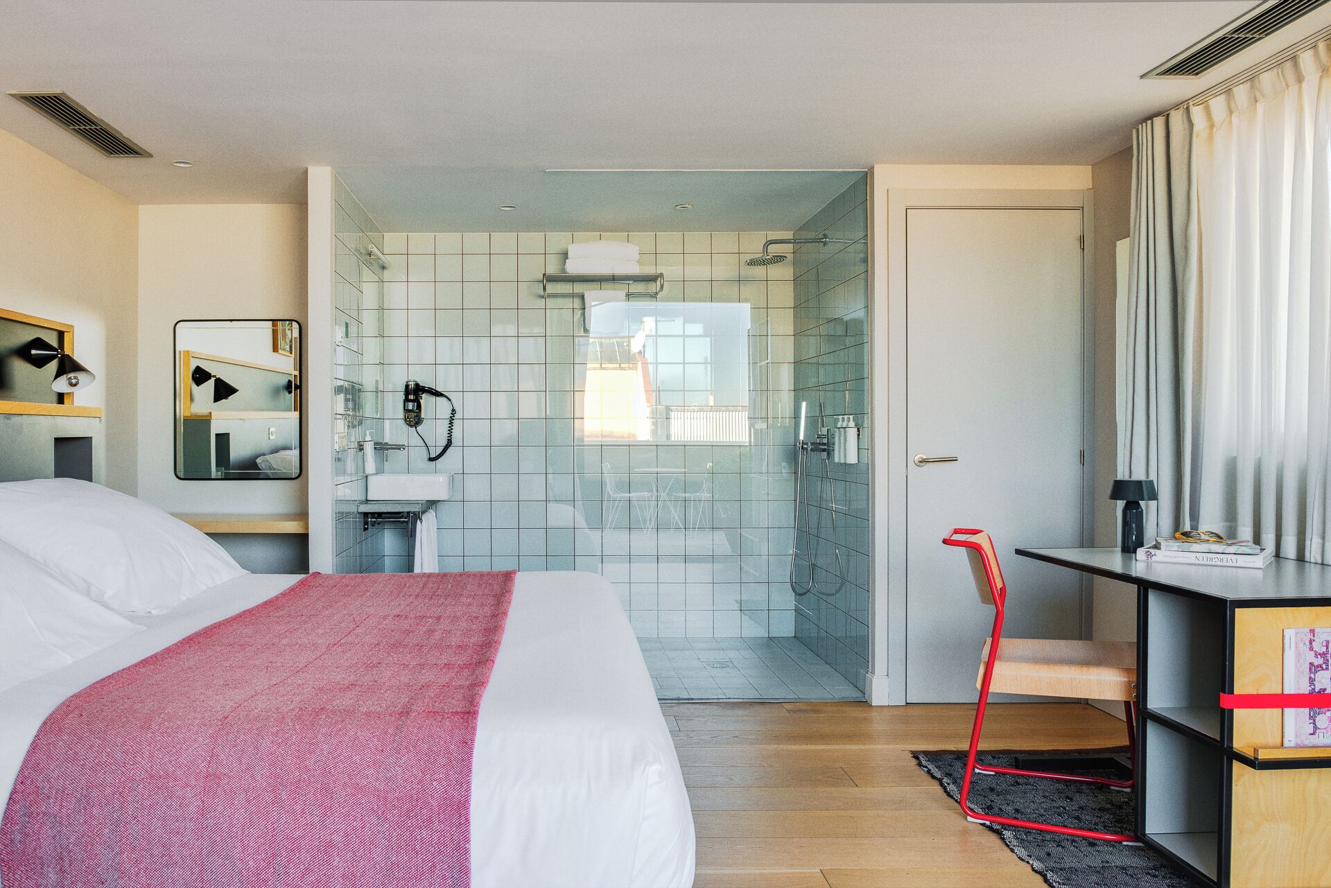 Quarto de hotel com cama, escrivaninha, cadeira, área de chuveiro e banheiro.