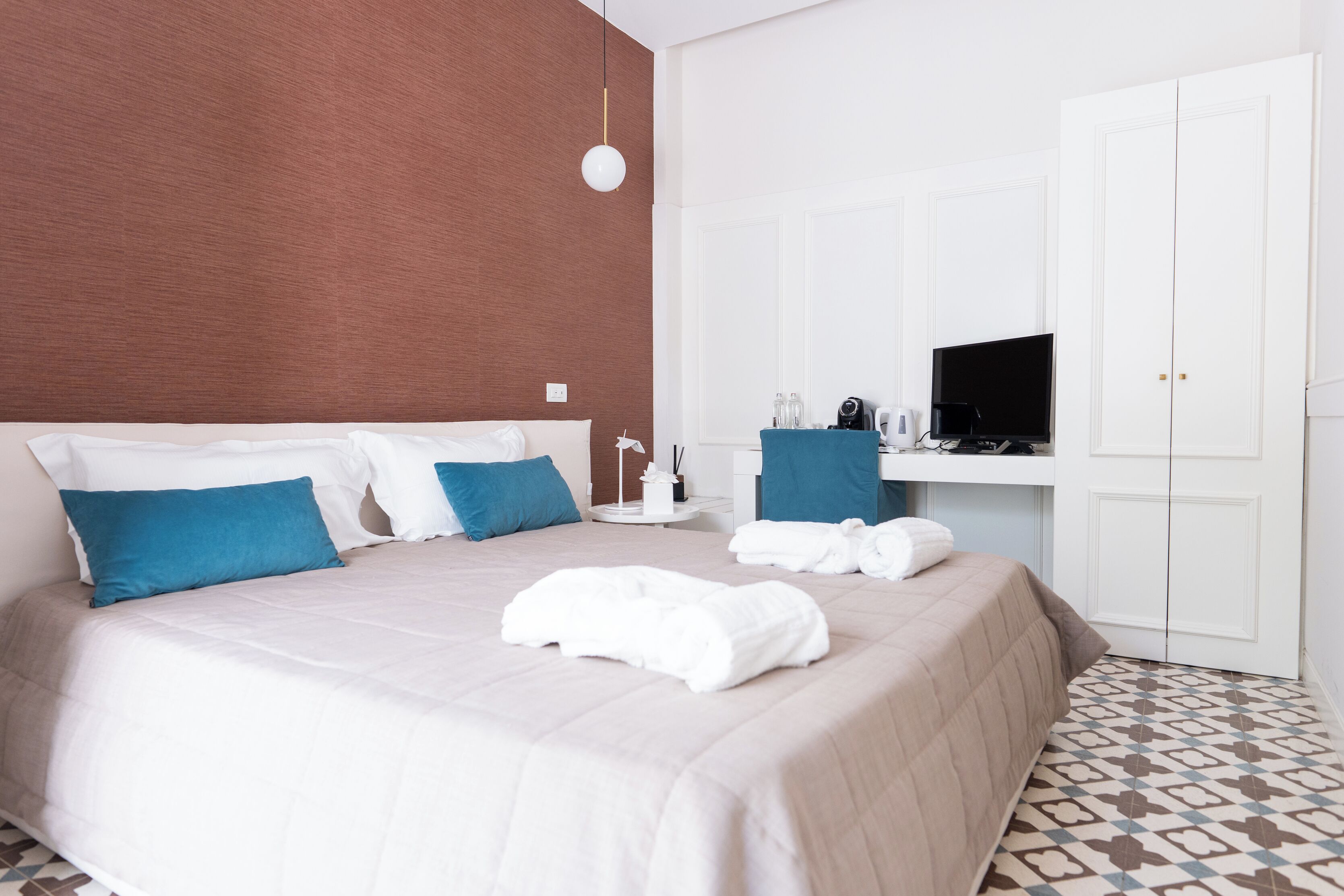 Cama bem arrumada com almofadas azuis e brancas, mesa de cabeceira branca com abajur, televisão e guarda-roupa em quarto de hotel.
