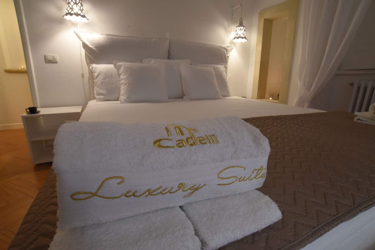 Cama bem arrumada com lençóis brancos e uma toalha com a inscrição 'Cadelli Luxury Suites'.