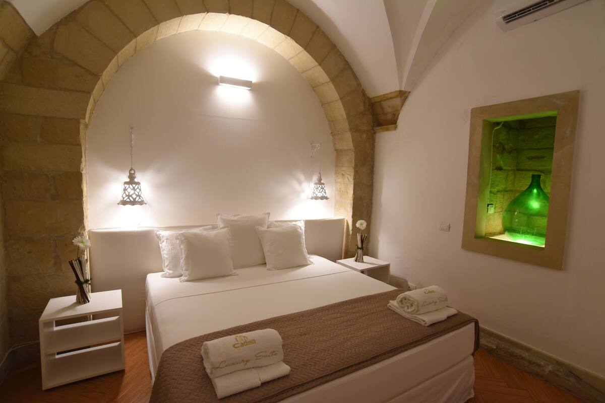 Quarto de hotel com uma cama grande, duas mesas de cabeceira, um criado-mudo, um vaso decorativo e uma garrafa iluminada por luz verde na parede.