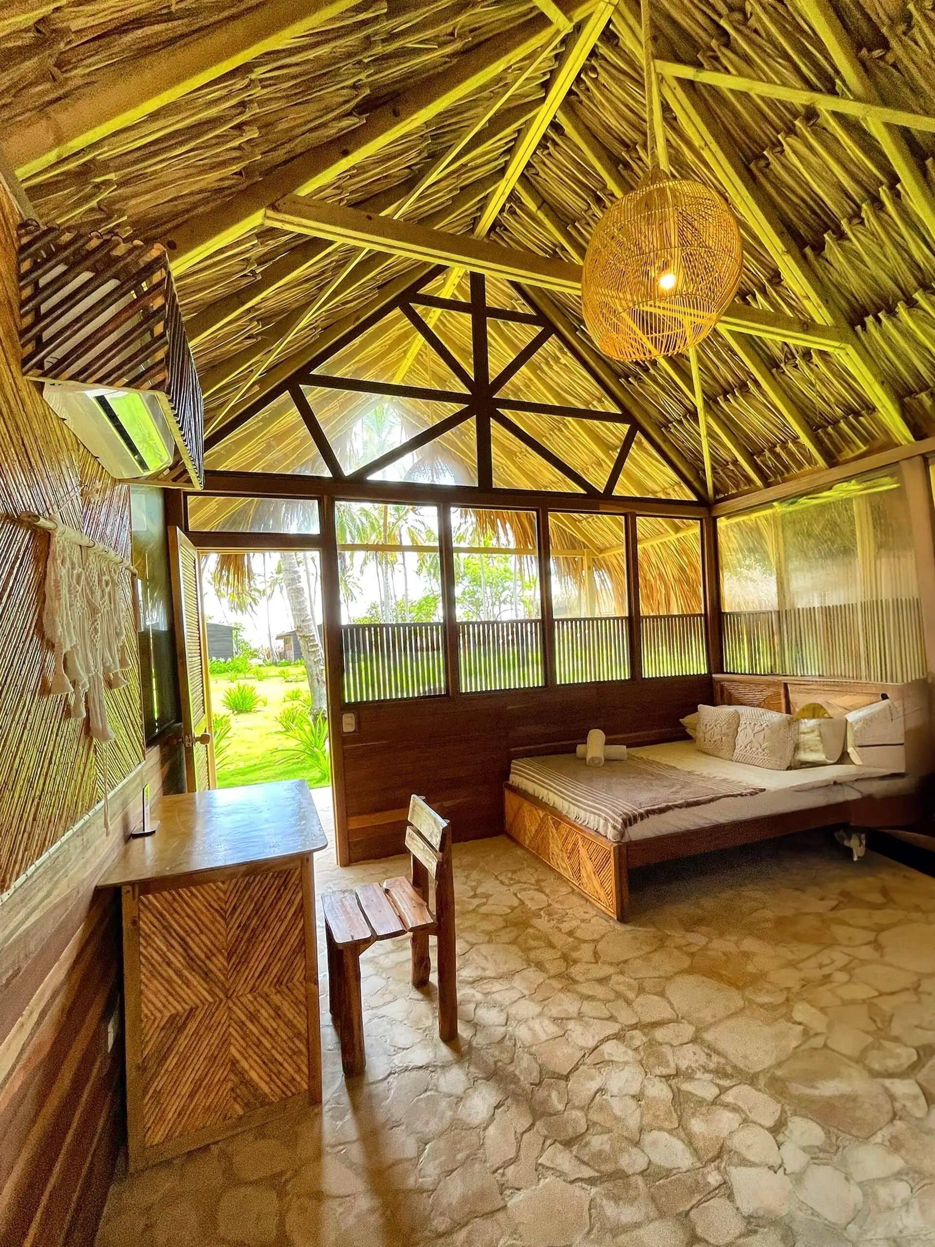 Um quarto com telhado de palha, uma cama com almofadas, um banco de madeira e uma mesa.
