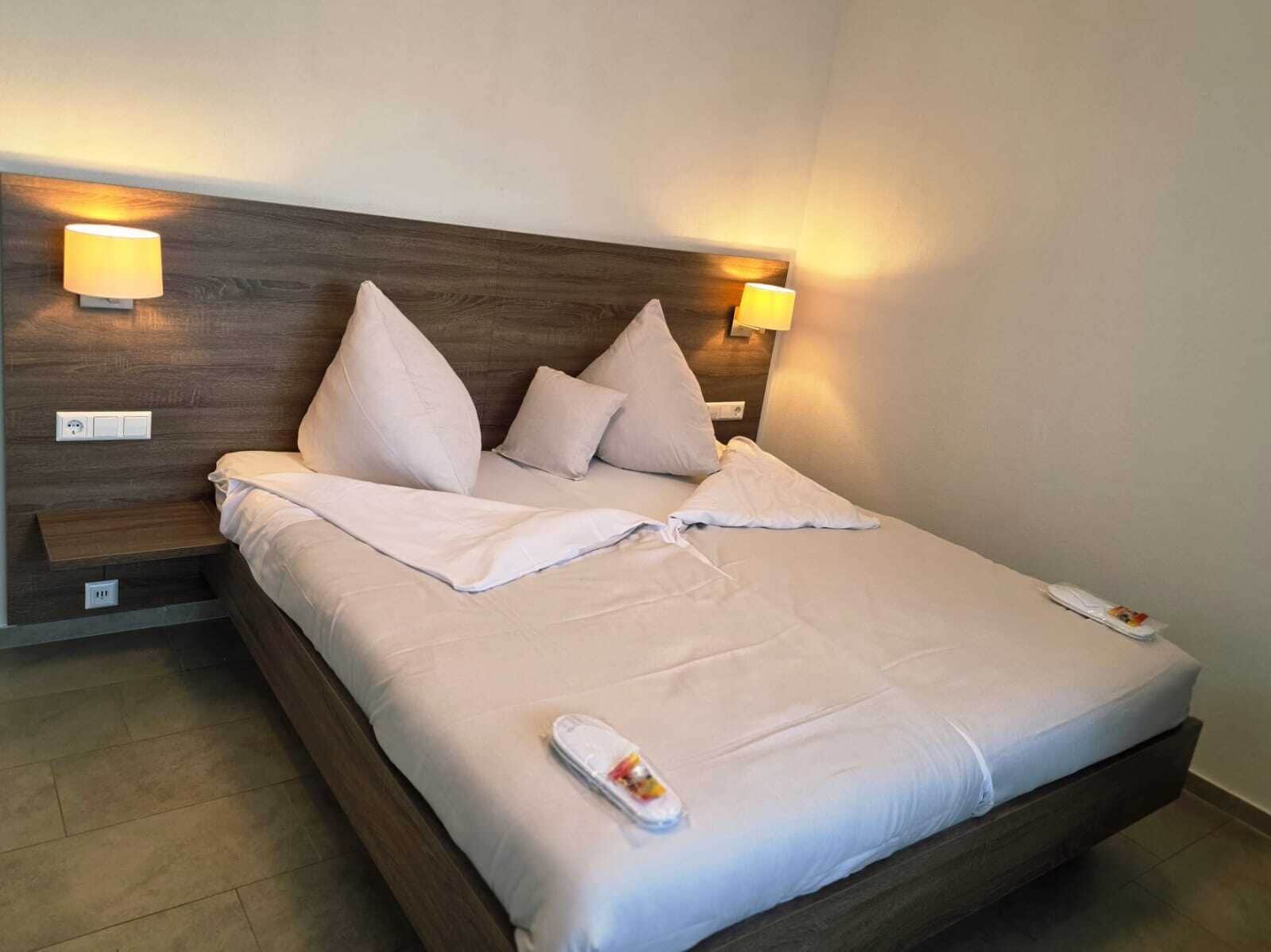 Cama bem arrumada com almofadas, mesa de cabeceira e duas luminárias fixadas na parede.