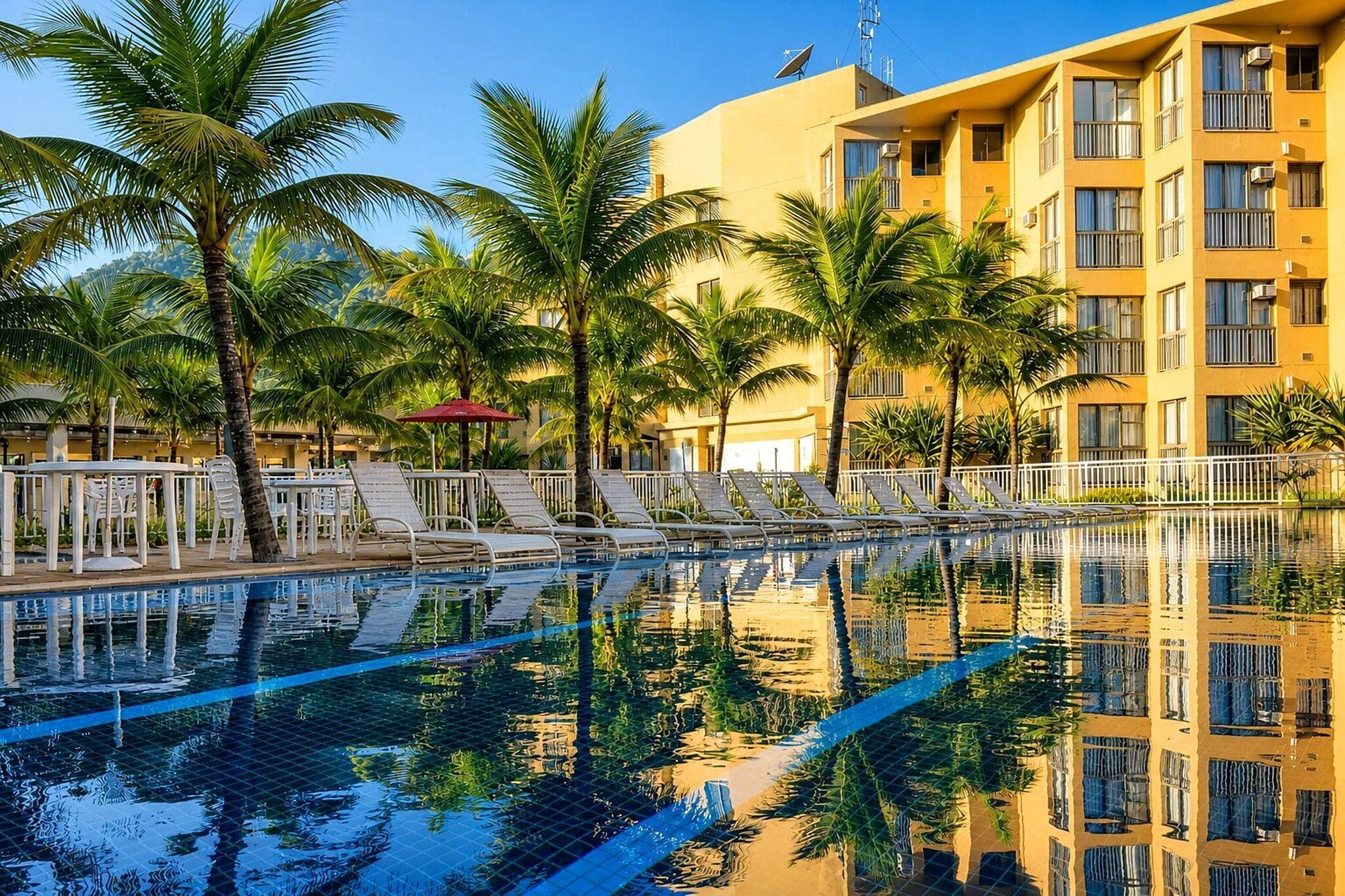 Piscina com espreguiçadeiras e palmeiras, rodeada por um edifício com v árias varandas.