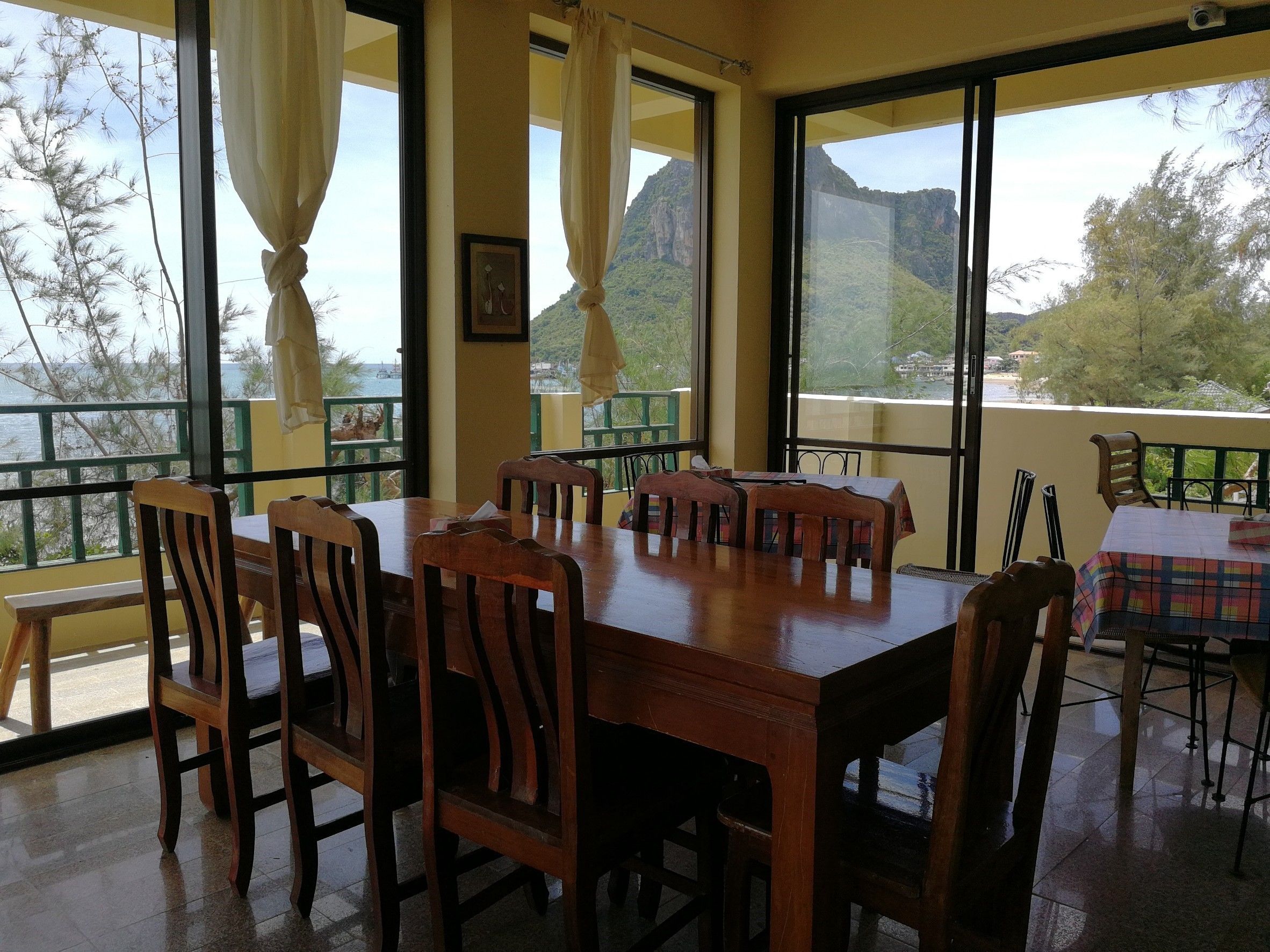 Uma sala de jantar com mesa e cadeiras de madeira, janelas amplas com cortinas e vista para montanhas e árvores.