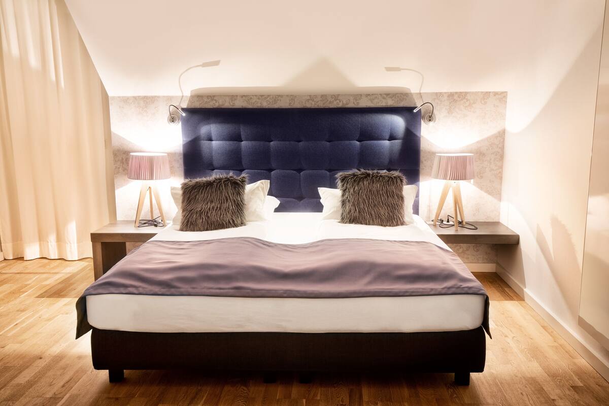 Quarto moderno com cabeceira estofada, cama grande com colcha roxa, duas mesas de cabeceira com luminárias e piso de madeira.